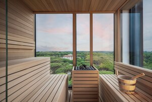 Um deck de madeira com sauna integrada, um balde de madeira e vista para uma paisagem verde através de grandes janelas.