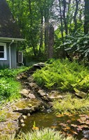 Um jardim tranquilo com um pequeno riacho, vegetação exuberante e uma casa com varanda.
