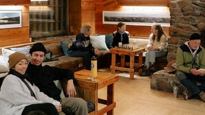 Um interior de madeira acolhedor, com lareira, uma mesa de xadrez e um grupo de pessoas sentadas.