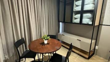 Quarto com mesa redonda de madeira, quatro cadeiras pretas, planta em vaso, banco embutido com espaço de arrumação e uma janela grande com persianas.