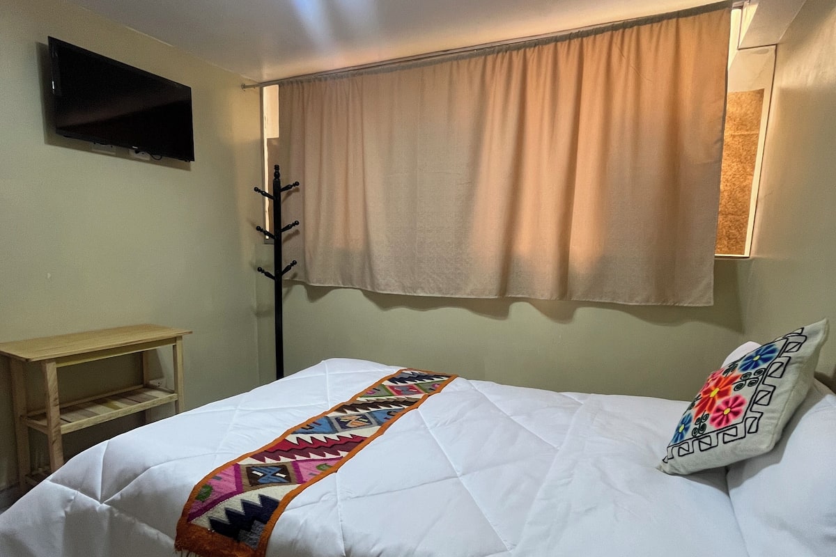 Quarto com cama, televisão fixada na parede, janela com cortina e mesa de cabeceira.