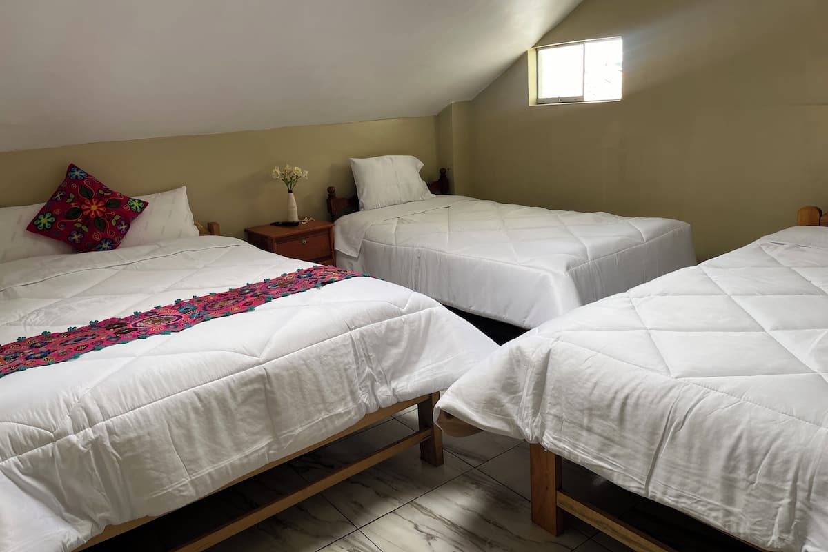 Quarto com duas camas, cada uma com roupa de cama branca e manta colorida. Há uma mesa de cabeceira com um vaso de flores e um abajur.