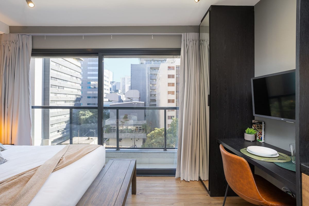Quarto de hotel com cama, escrivaninha com cadeira, televisão e uma janela grande com cortinas.