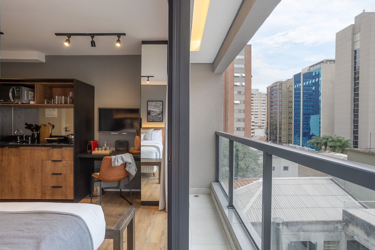 Quarto de hotel moderno com cozinha compacta, cama, escrivaninha e varanda com vista para a cidade.