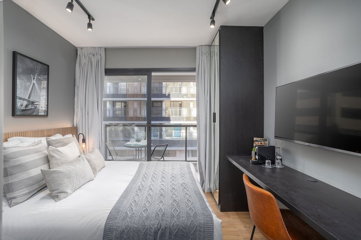Quarto de hotel moderno com cama, secretária, televisão de tela plana, janela grande com cortinas e varanda.