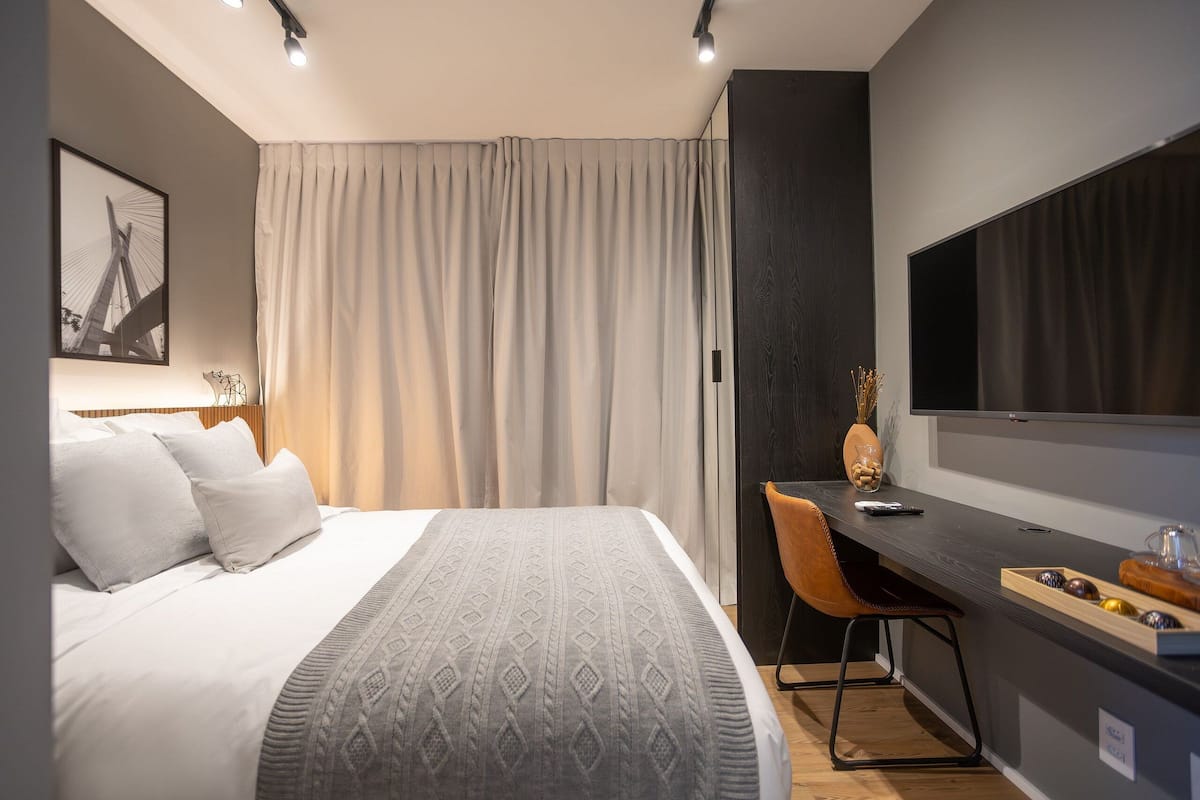 Quarto de hotel moderno com cama, escrivaninha, cadeira, TV de tela plana e uma janela grande com cortinas.