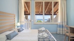 Quarto com cama, almofadas, toalha enrolada, cadeira, janela com cortinas e vista para o exterior.