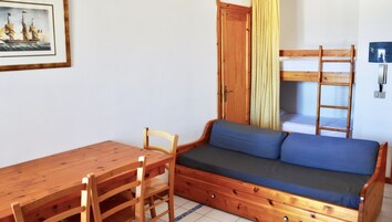 Quarto compacto com beliche de madeira, sofá-cama com gavetas de armazenamento, mesa de madeira com cadeiras e um quadro emoldurado na parede.