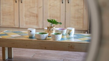 Uma mesa de madeira com toalha quadriculada, rodeada por seis chávenas de cerâmica e uma cesta de vime com uma planta.