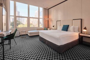 Quarto de hotel com uma cama grande, uma escrivaninha, uma cadeira, uma mesinha e um radiador.