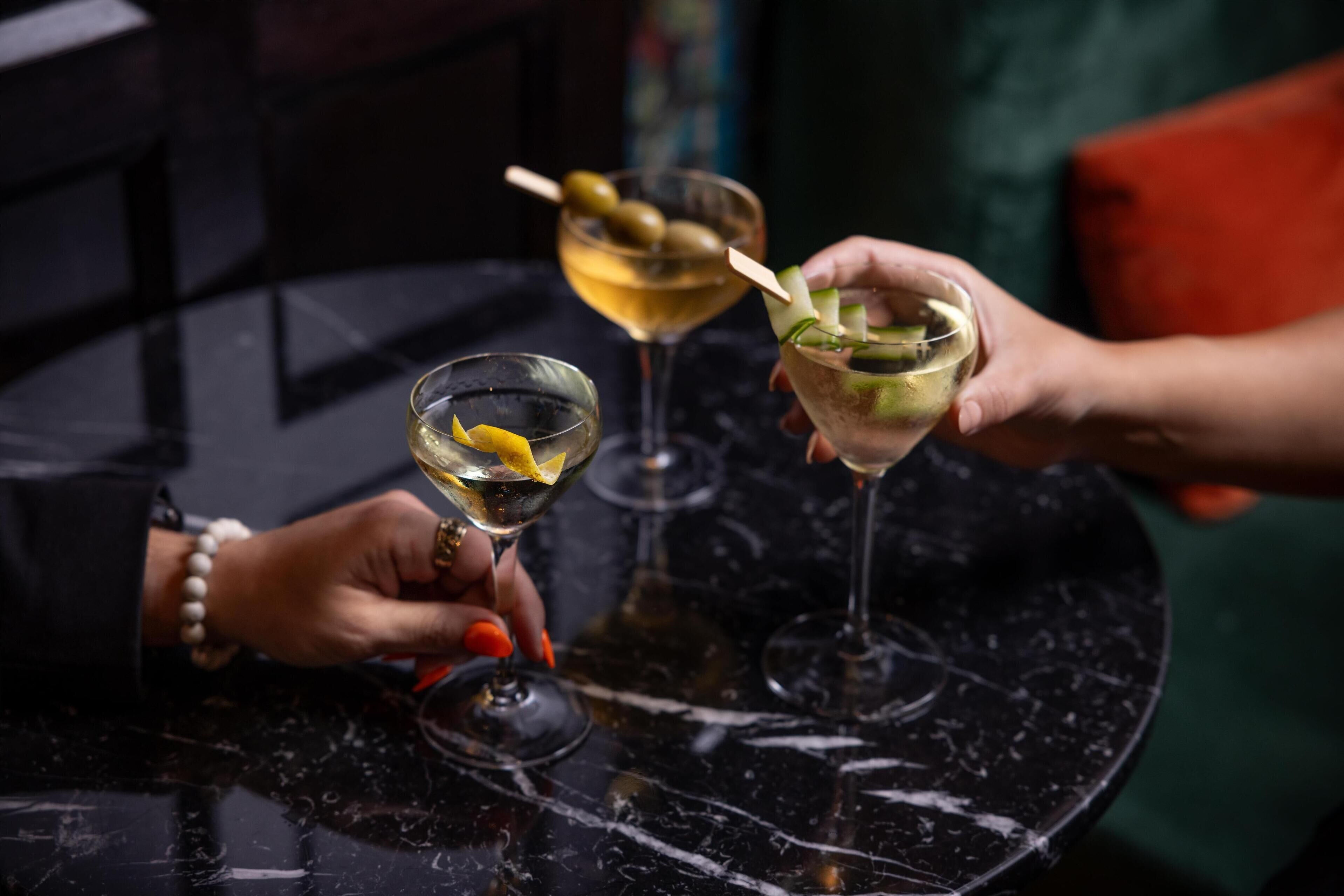 Uma mesa de mármore com três copos de cocktails: um com uma fatia de limão e pepino, outro com azeitonas, e um terceiro com pepino.