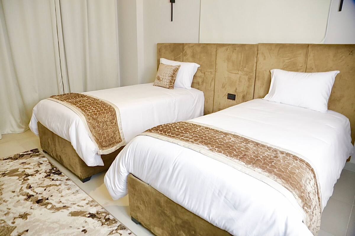 Duas camas com roupa de cama branca e castanha, uma cabeceira de madeira e um tapete estampado.