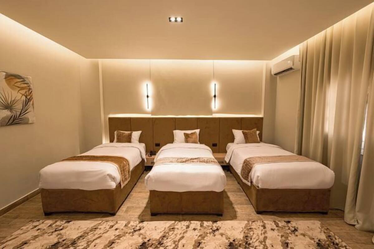 Quarto de hotel com três camas, uma cabeceira grande, uma luminária no teto e uma janela com cortinas.