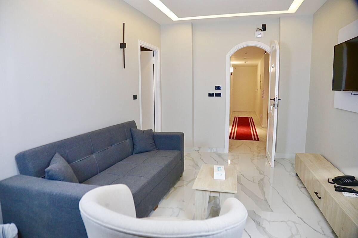 Uma sala de estar moderna com um sofá cinzento, uma poltrona branca, uma pequena mesa de centro de madeira e uma TV fixada na parede.