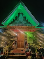 Entrada de hotel com uma placa iluminada em verde com a inscrição "HOTEL MOUNTAIN" e estilo arquitetônico tradicional.