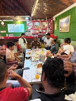 Um grupo de pessoas sentadas à mesa, com diversos pratos e bebidas, em um espaço com paredes verdes e teto de madeira.