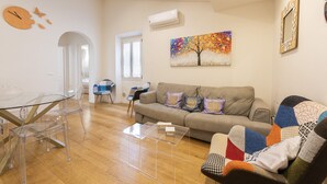 Uma sala de estar com uma mesa de centro de vidro, um sofá com almofadas estampadas, um quadro de uma árvore e um relógio na parede.