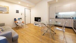 Um espaço de estar moderno com mesa de jantar de vidro, cadeiras transparentes, sofá cinza, televisão de tela plana e área de cozinha com armários e bancada.