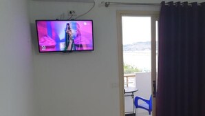 Quarto com televisão de tela plana na parede, janela com cortinas, mesa pequena com uma cadeira e vista para o exterior.