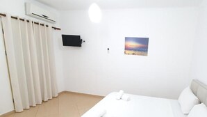 Quarto moderno e limpo, com cama, cortinas brancas, televisão na parede e um quadro.