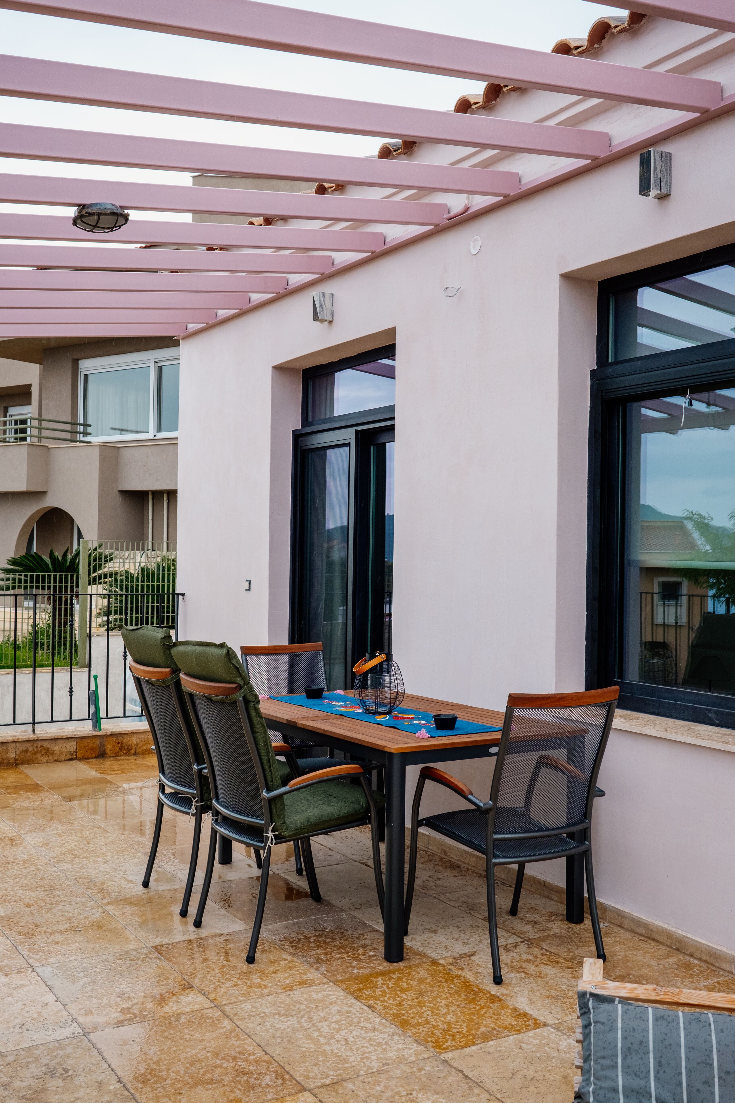 Um pátio externo com mesa e cadeiras, uma pérgula e vista para um edifício com grandes janelas.