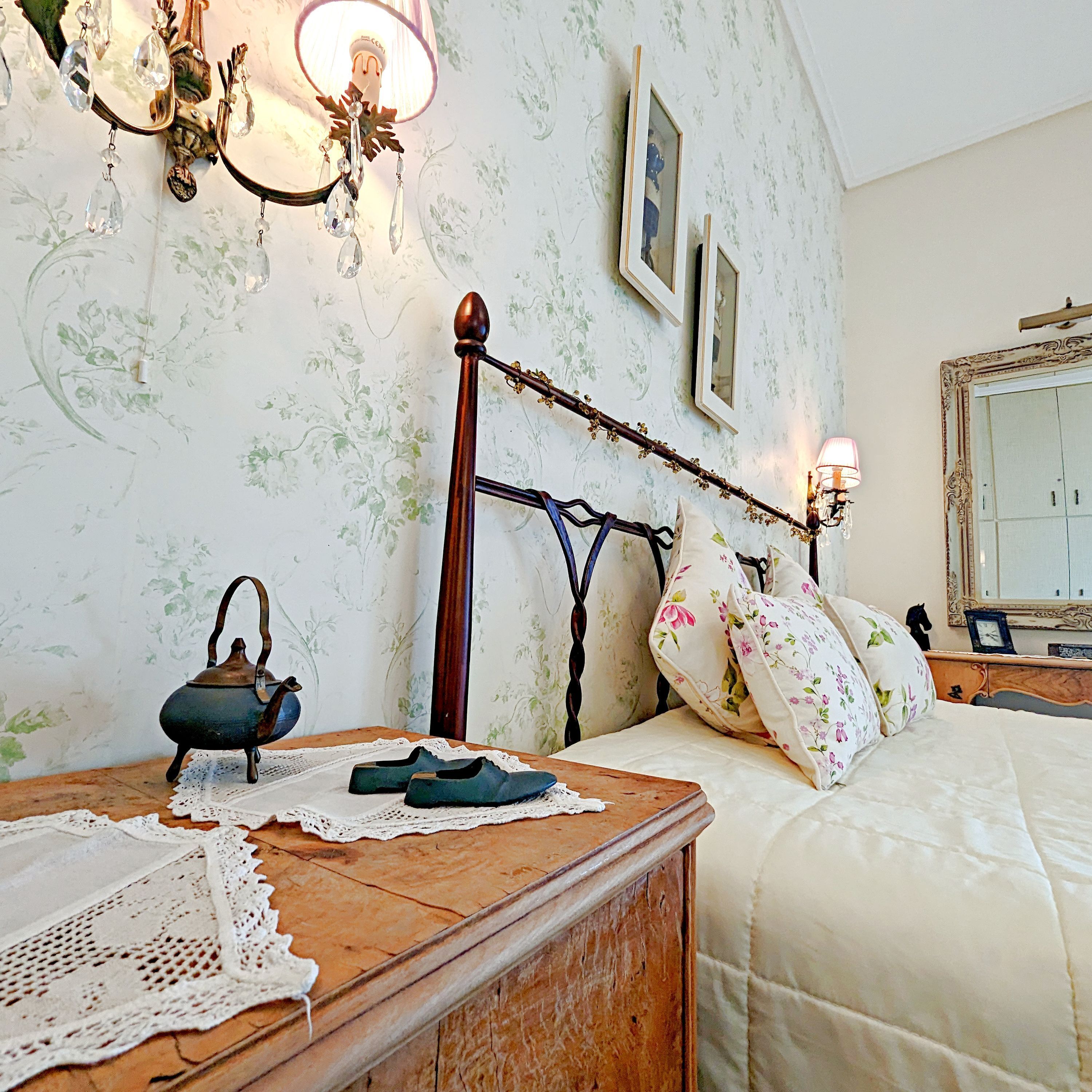 Quarto com papel de parede floral, estrutura de cama de madeira, mesa de cabeceira com bule e espelho com moldura decorativa.