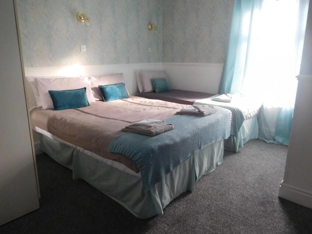 Quarto com duas camas, cada uma com edredom e travesseiros, uma mesinha de cabeceira e uma janela com cortinas.