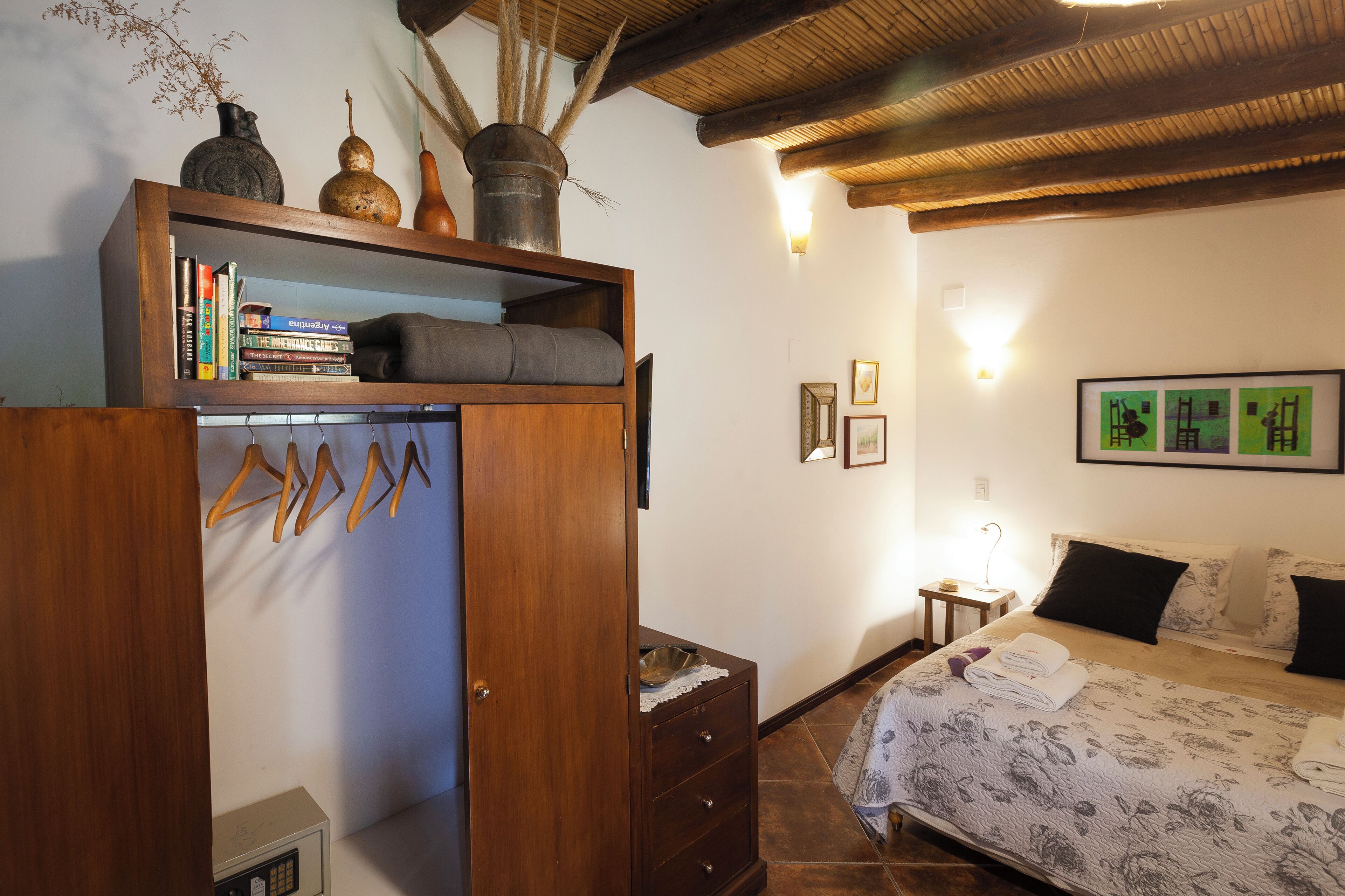 Quarto com cama, guarda-roupa de madeira, mesa de cabeceira e prateleira na parede com livros e objetos decorativos.