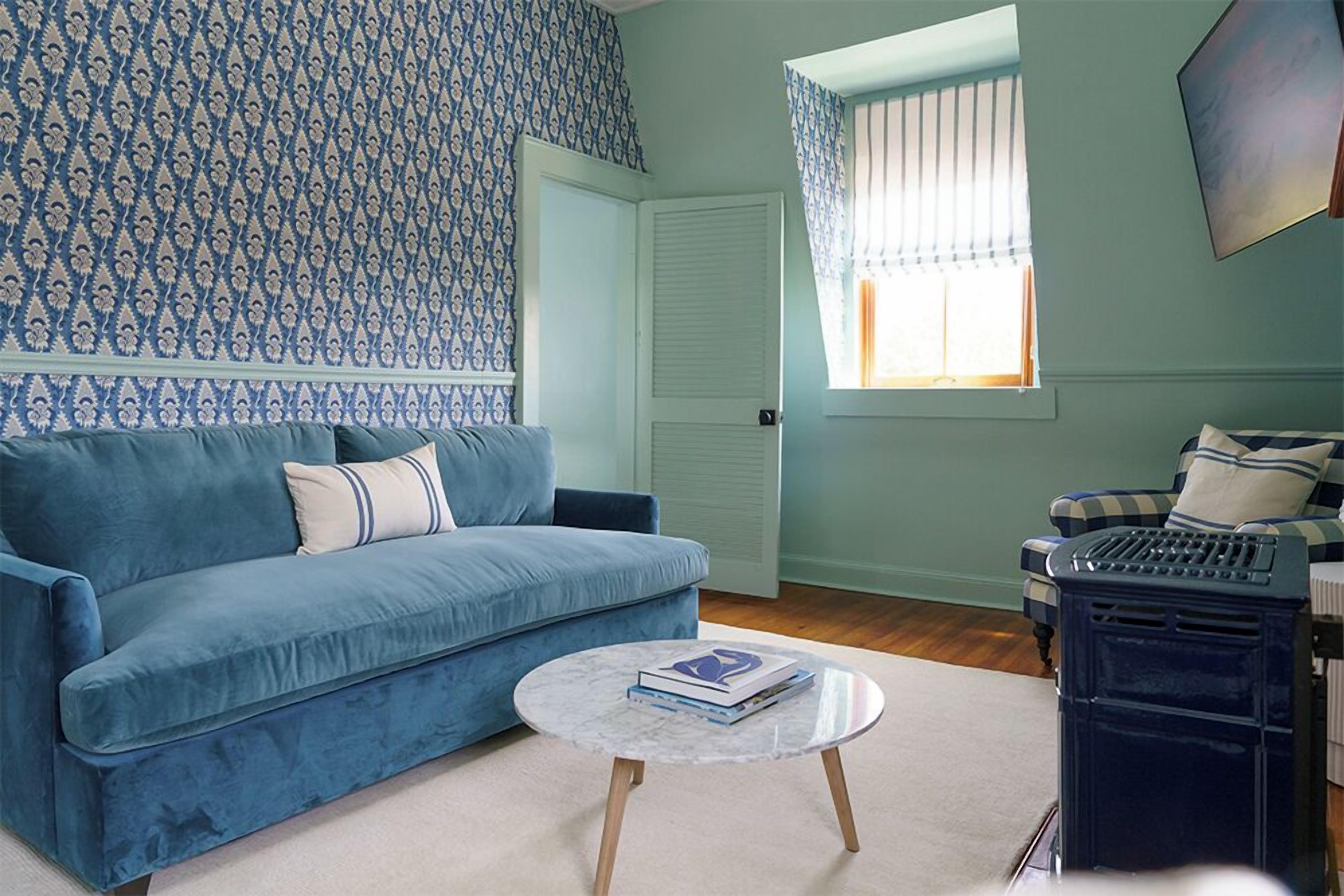 Quarto com sofá azul, uma pequena mesa redonda com livros, papel de parede estampado, uma janela com persianas e uma cadeira com almofada.