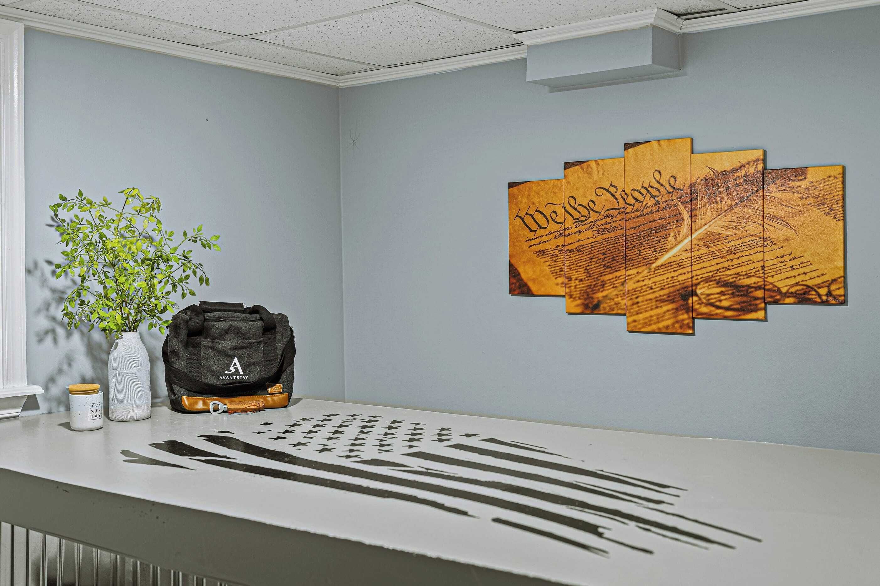 Um quarto com uma escrivaninha, um vaso com planta, uma mochila preta e um quadro de parede com o cartaz 'We the People'.