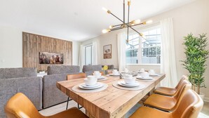 Uma sala de jantar moderna com uma mesa de madeira posta para quatro pessoas, rodeada por cadeiras de couro. Um sofá cinzento e uma pequena mesa de apoio com uma luminária são visíveis ao fundo.