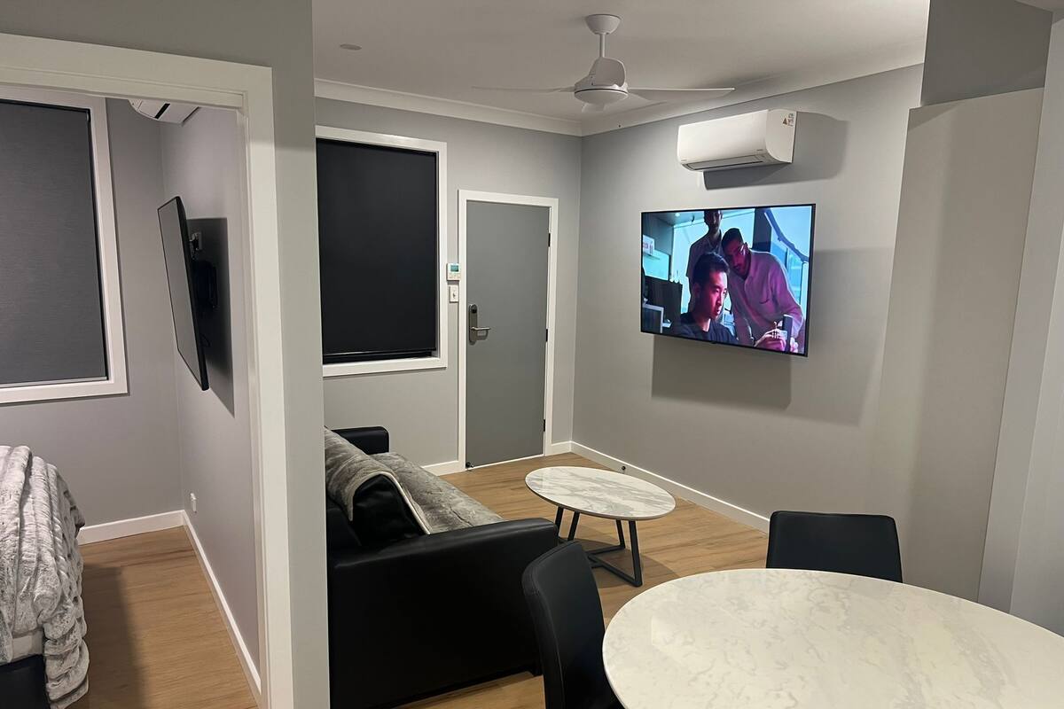 Uma sala de estar moderna com uma televisão de tela plana na parede, um sofá cinza, uma mesa de centro redonda e um ventilador de teto.