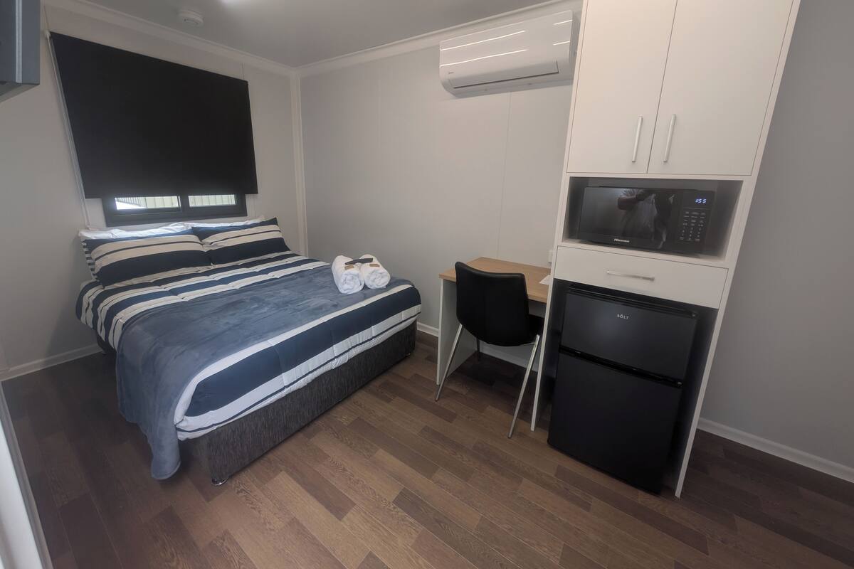 Quarto compacto com cama, escrivaninha, cozinha compacta e TV na parede.