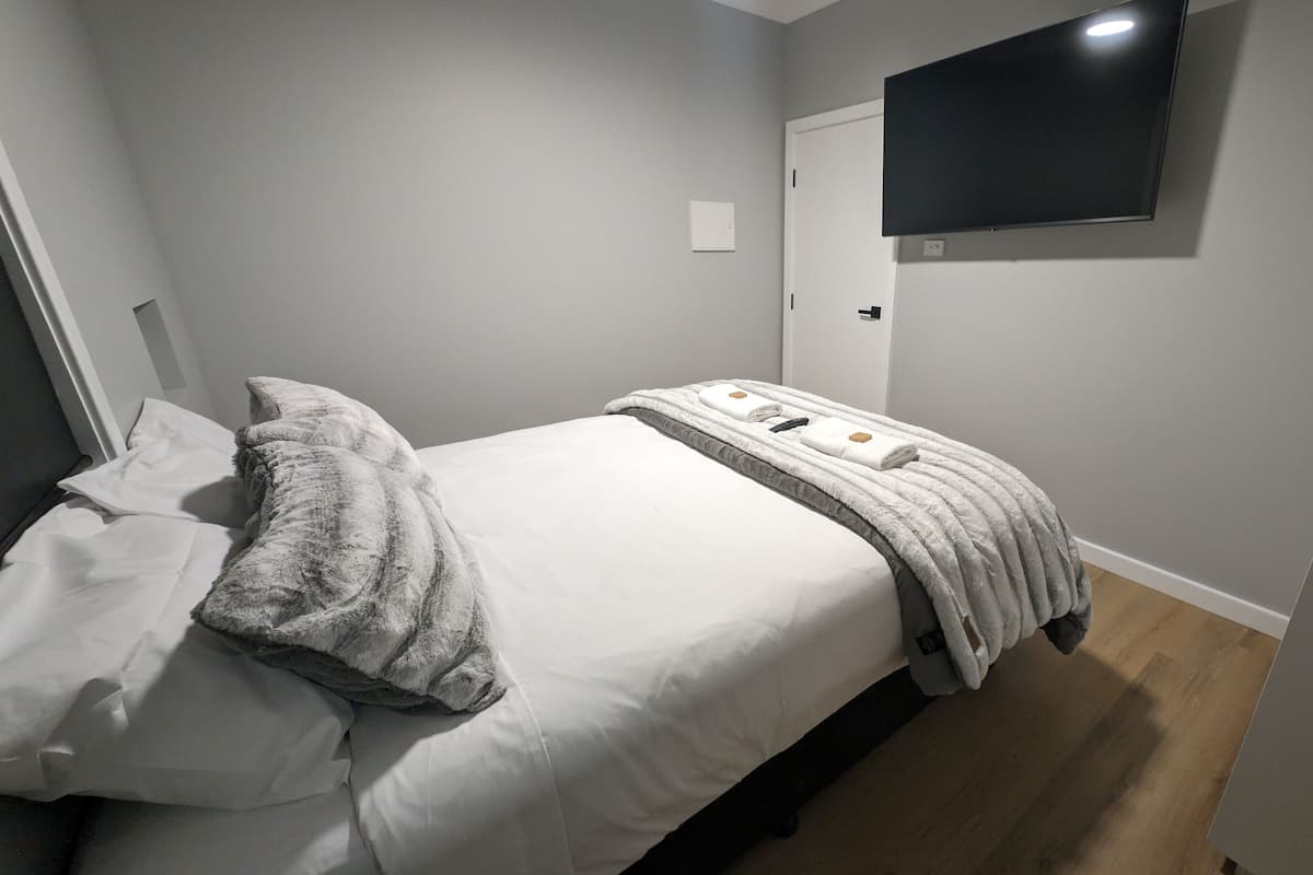 Uma cama bem arrumada com lençóis brancos e um cobertor fofinho, uma televisão de tela plana na parede e piso de madeira.