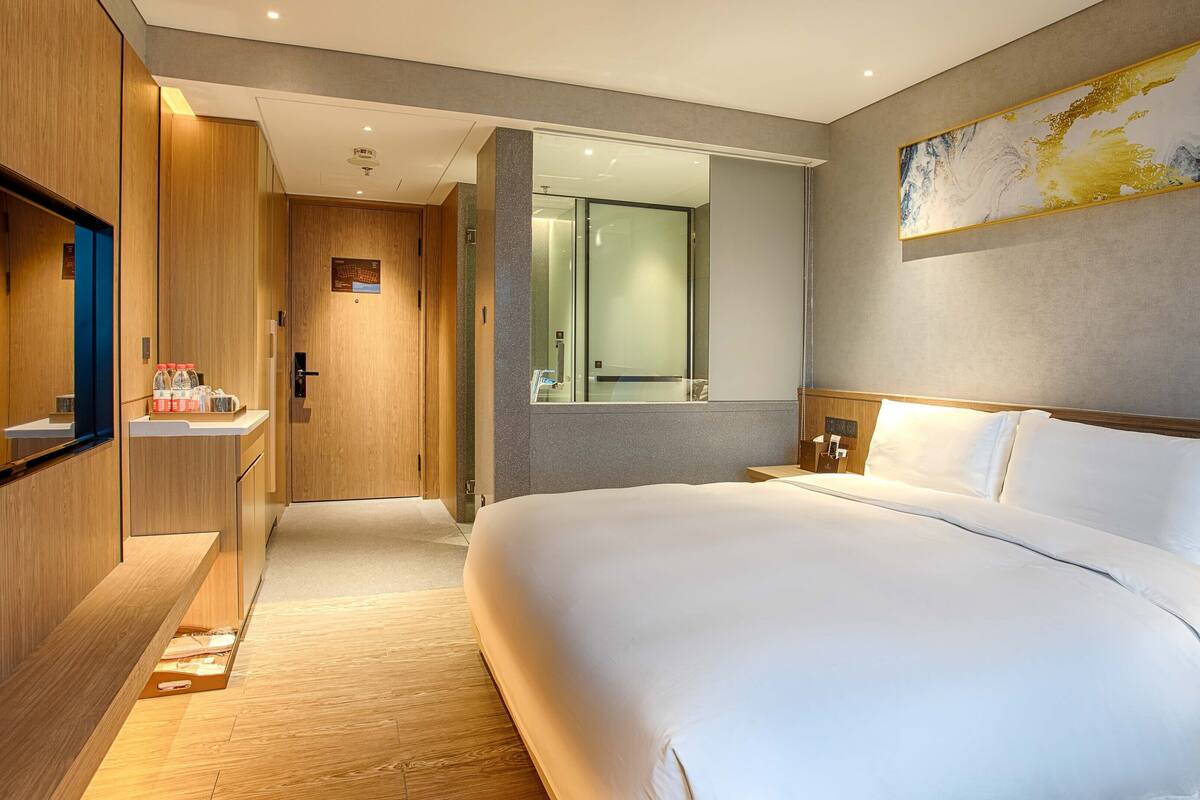 Quarto de hotel moderno com uma cama grande, televisão de tela plana na parede, guarda-roupa de madeira e banheiro visível através de uma porta de vidro.