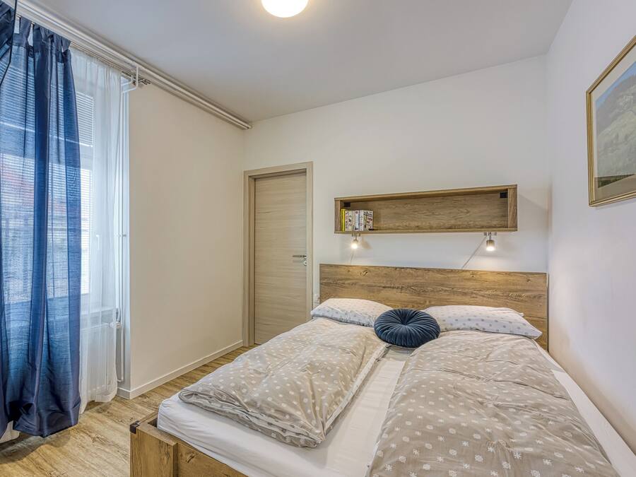 Quarto com cabeceira de madeira, mesa de cabeceira, cama com roupa de cama estampada, janela com cortinas leves e prateleira com livros.