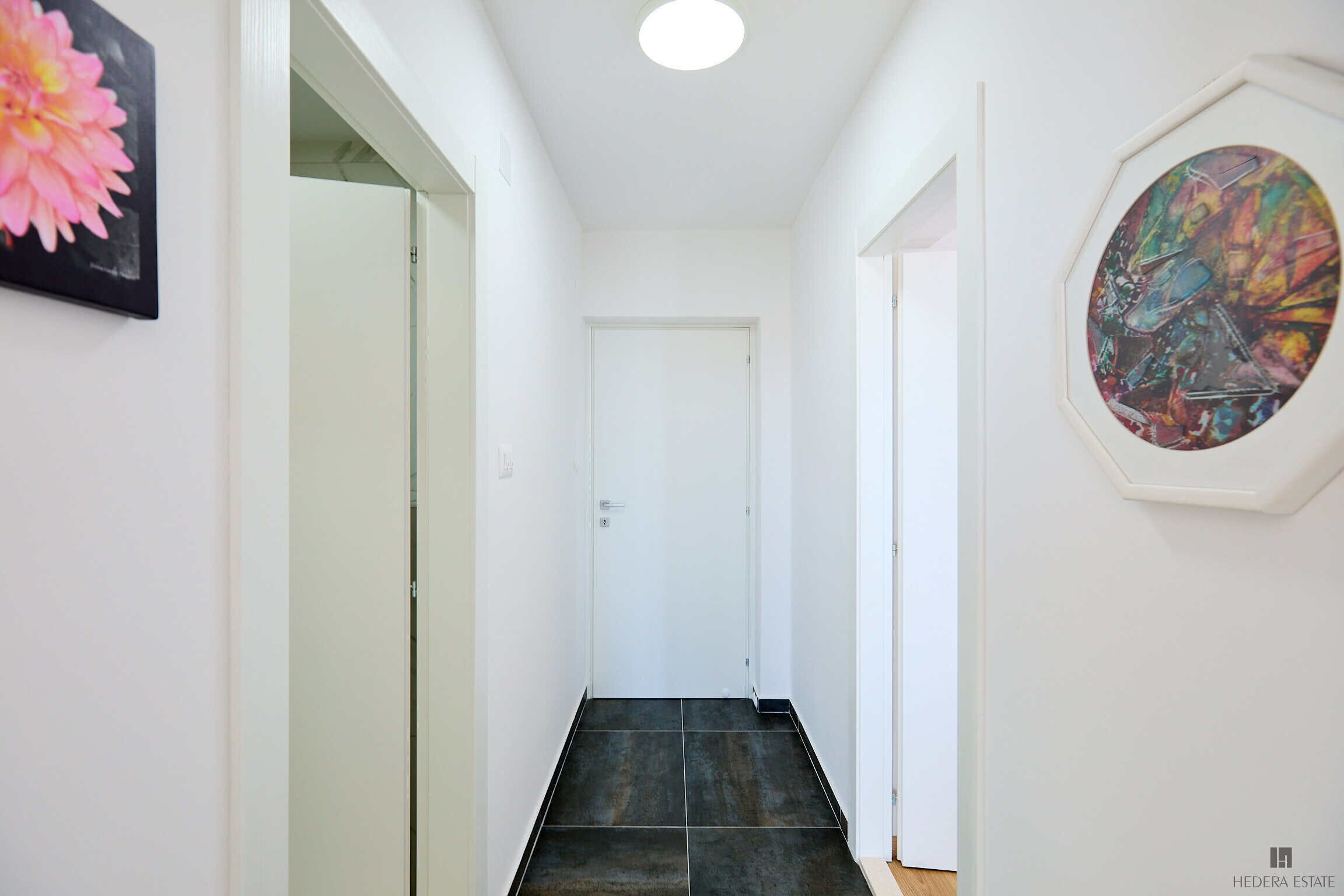 Um corredor com uma porta branca no fim, chão de piso preto e branco e uma pintura na parede direita.