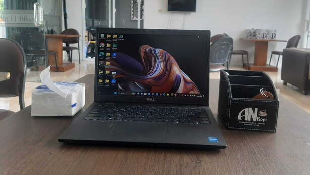 Um laptop sobre uma mesa de madeira num quarto com cadeiras, um vidro e uma televisão na parede.