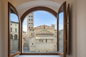 Visão através de uma janela em arco de um edifício histórico, com uma torre de sino, um pátio e arquitetura circundante.