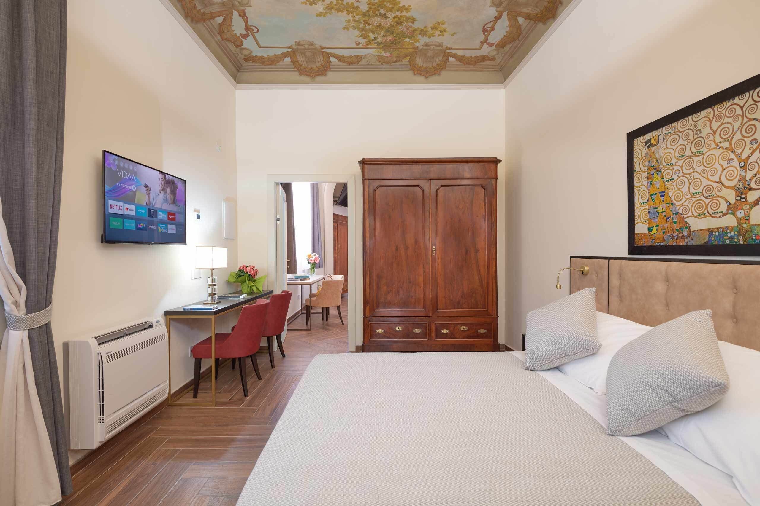 Quarto de hotel com cama, escrivaninha, guarda-roupa e televisão exibindo um programa.