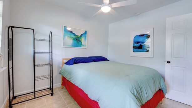Quarto com cama, ventilador de teto, estante de metal e duas pinturas abstratas nas paredes.