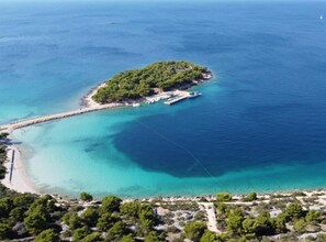Vista aérea de uma pequena ilha com mar azul transparente, rodeada por um litoral rochoso e alguns edifícios.
