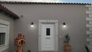 Uma casa com porta branca, duas luzes fixadas na parede, um comedouro para pássaros e um vaso de planta.
