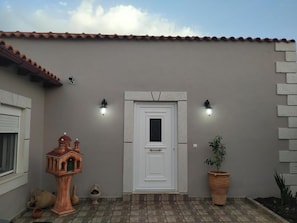 Uma casa com porta branca, duas luzes fixadas na parede, um comedouro para pássaros e um vaso de planta.
