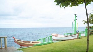 Vista costeira com área gramada, dois barcos, cerca verde e uma construção alta.
