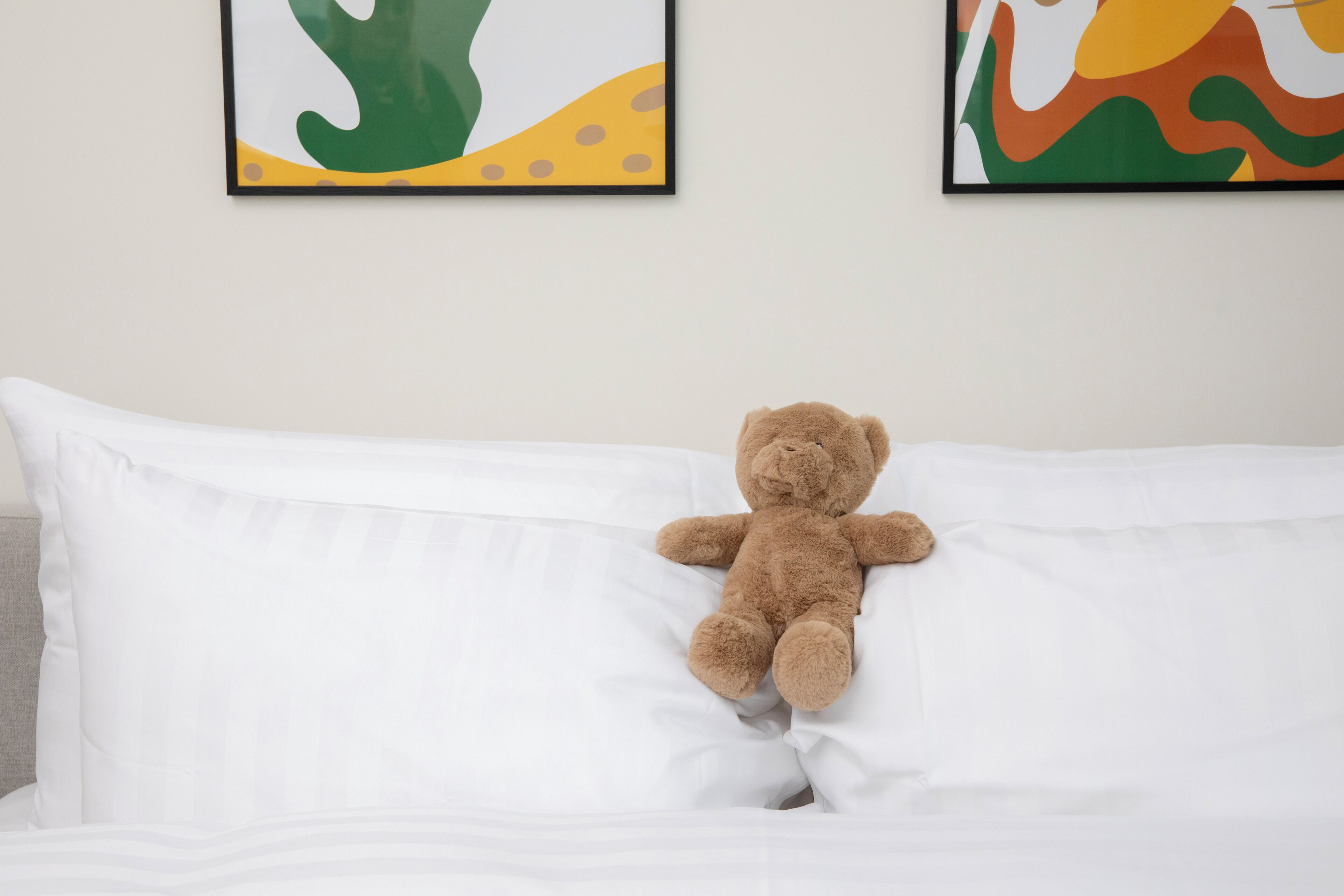 Uma cama com um ursinho de pelúcia no travesseiro, roupa de cama branca e duas pinturas abstratas na parede.