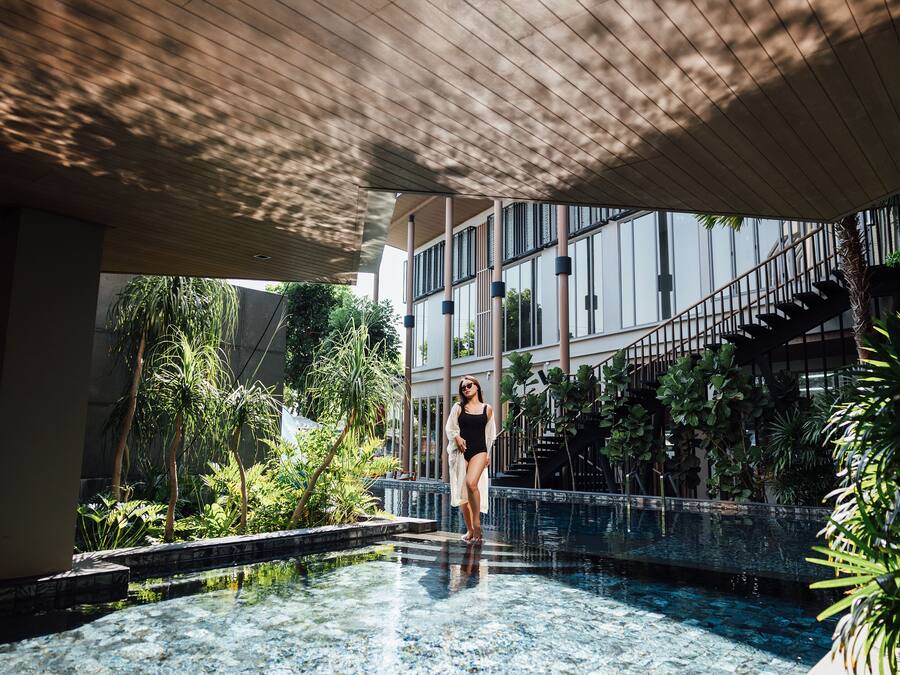 Uma mulher de fato de banho preto está na beira de uma piscina com teto de madeira e uma escadaria ao fundo.