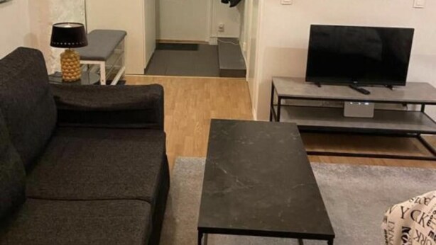 Uma sala de estar com um sofá cinza escuro, uma pequena mesa de centro preta, uma televisão em um suporte e teto com vigas de madeira.