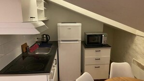 Cozinha compacta com micro-ondas, mini-geladeira, pia e fogão. Armários brancos e mesa de madeira com cadeiras.