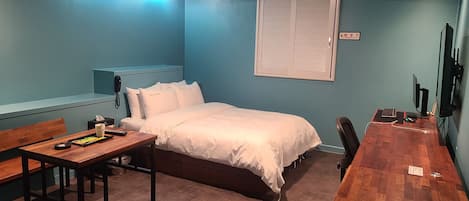 Quarto com cama, escrivaninha, cadeira, televisão e janela com persianas.